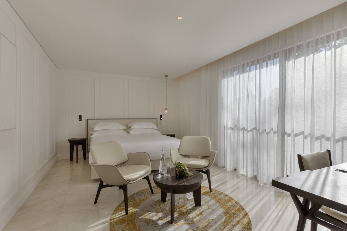 Quarto de hotel moderno com uma cama grande, duas cadeiras, uma mesa pequena e vista da cidade através de cortinas leves.