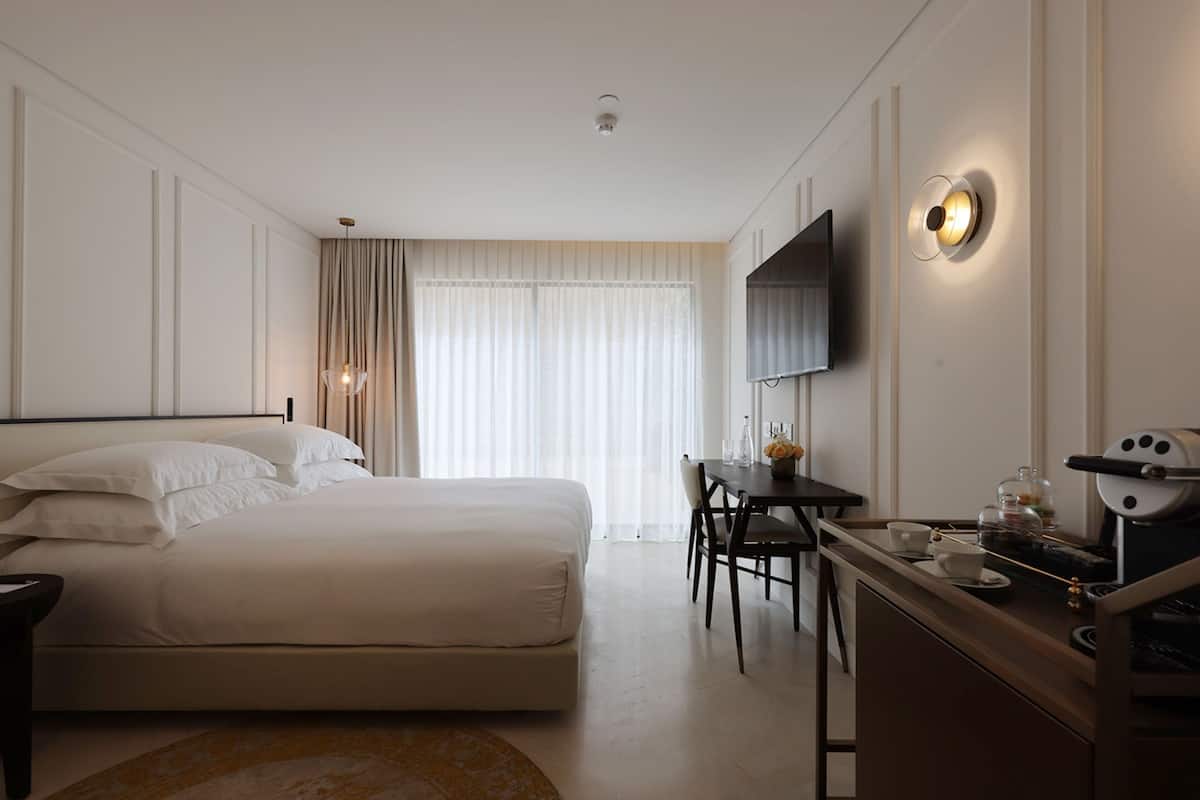 Quarto de hotel com cama, escrivaninha com cafeteira, TV na parede e janela com cortinas.