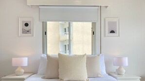 Cama arrumada com roupa de cama e almofadas brancas, duas luminárias de cabeceira e uma janela com vista para uma varanda.