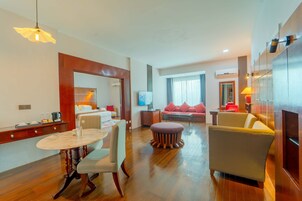 Quarto de hotel com área de refeições, sala de estar com sofá e televisão, e um espelho grande.