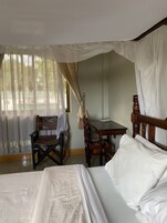 Quarto com cama, cadeira de madeira, mesa pequena e janela com cortinas finas.
