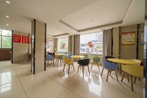 Um hall de hotel com balcão de recepção, área de estar com mesas e cadeiras, e grandes janelas.
