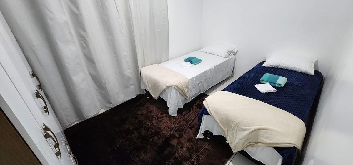 Quarto pequeno e compacto com duas camas de solteiro, carpete marrom escuro e cortinas brancas.