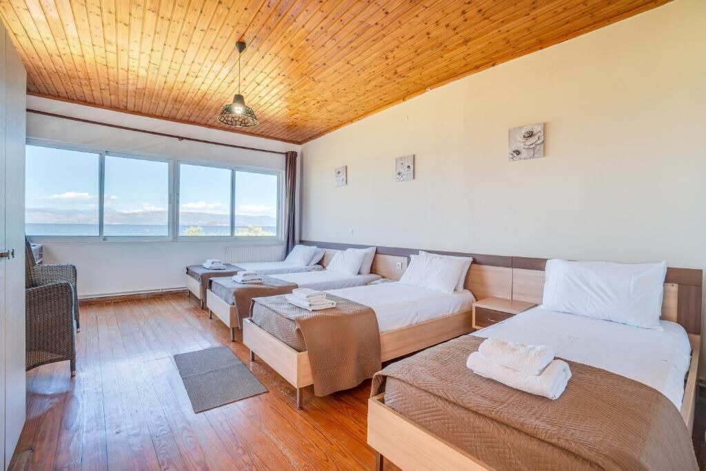 Quarto de hotel com três camas, teto de madeira, janelas amplas e vista para o mar.