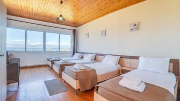 Quarto de hotel com três camas, teto de madeira, janelas amplas e vista para o mar.