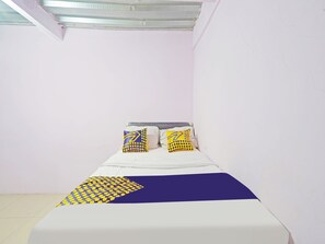 Quarto pequeno com uma cama de solteiro, colcha branca, duas almofadas com estampas em amarelo e azul, e um cobertor azul.