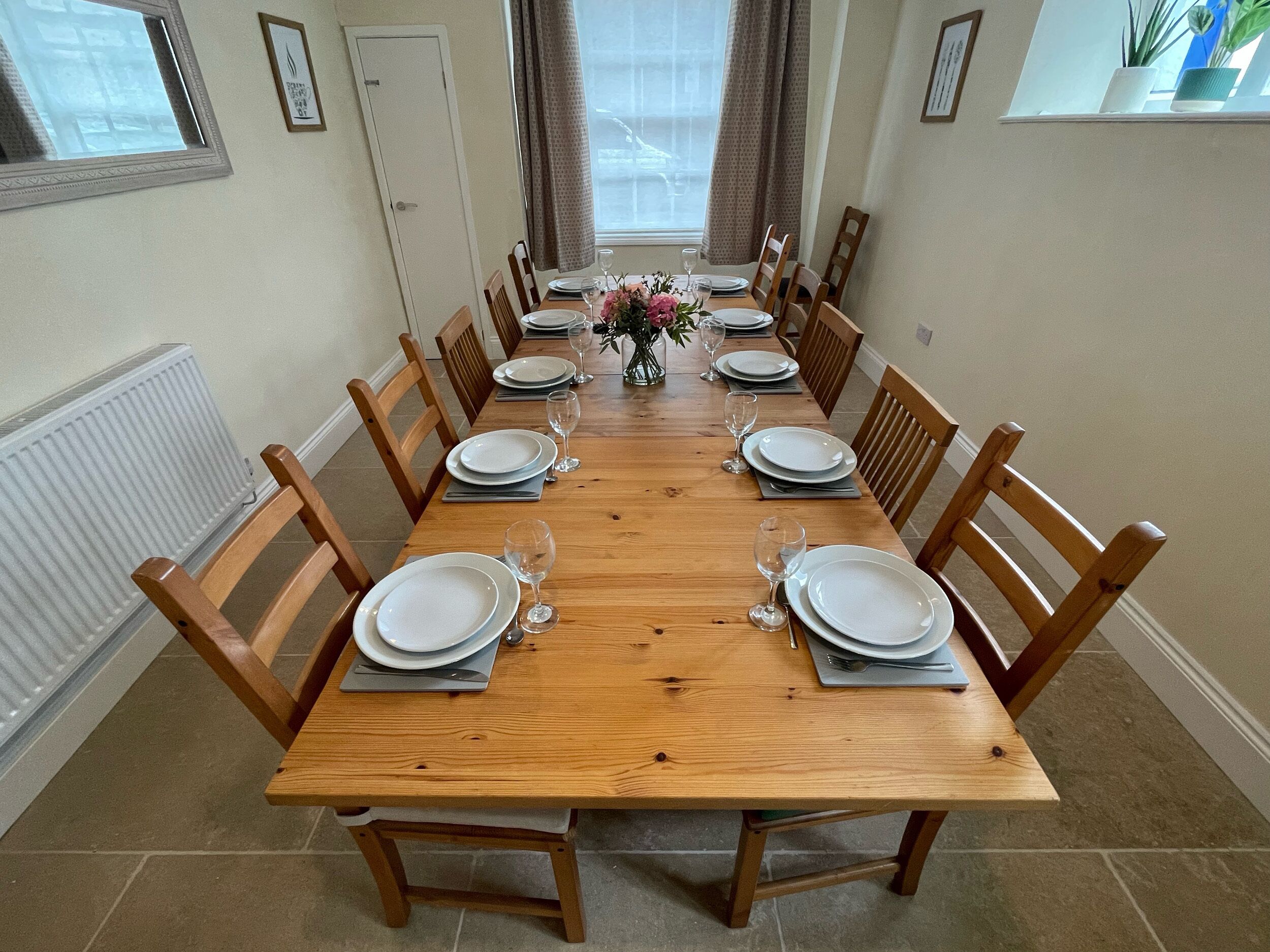 Uma sala de jantar com uma longa mesa de madeira posta para uma refeição, rodeada por cadeiras de madeira.