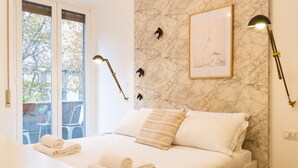 Um quarto com cama, duas luminárias de cabeceira, um quadro na parede e vista para uma varanda com cadeiras e planta em vaso.