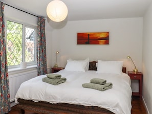 Cama bem arrumada com duas almofadas e duas toalhas dobradas, pintura de um pôr do sol, janela com cortinas e uma luminária em cada lado da cama.