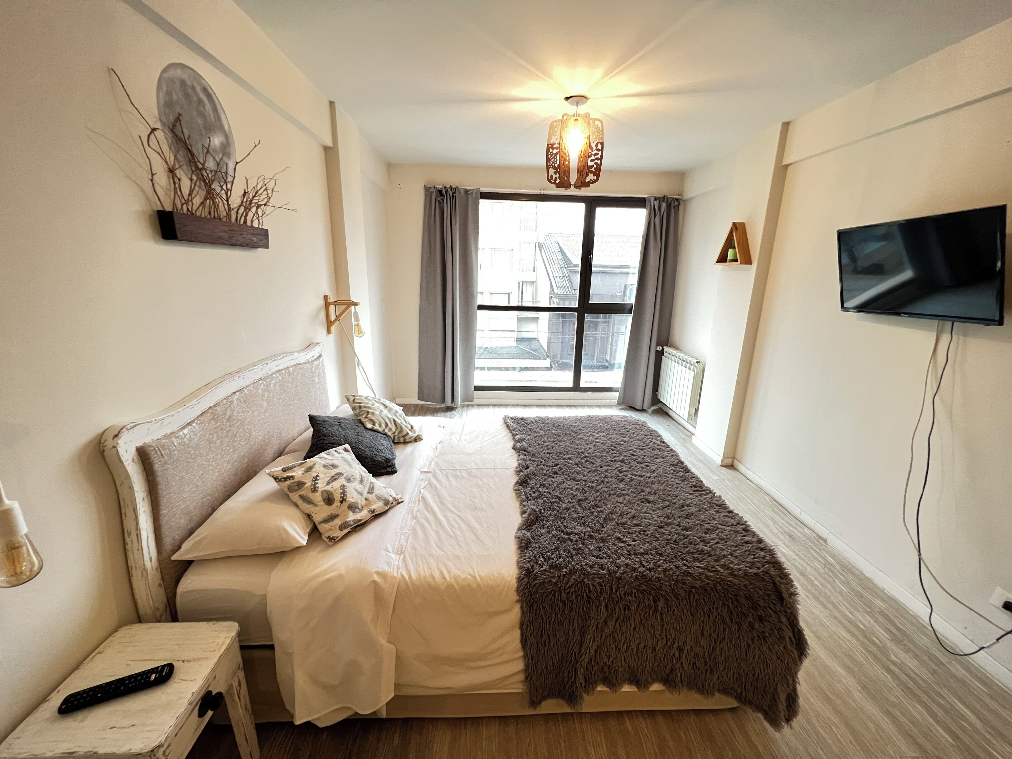Quarto com cama, televisão, janela com cortinas, ventoinha no teto e luminária fixada na parede.