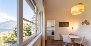 Uma cozinha moderna com armários brancos, uma área de jantar com mesa e cadeiras redondas, e vista para um corpo de água e montanhas através de grandes janelas.