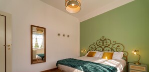 Quarto com cama, mesa de cabeceira, abajur, espelho e porta.