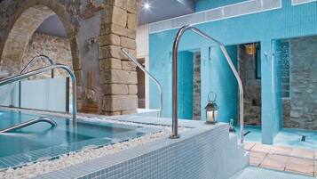 Uma piscina termal com arcos de pedra, paredes revestidas de azulejos e uma parede de pedra com um candeeiro.