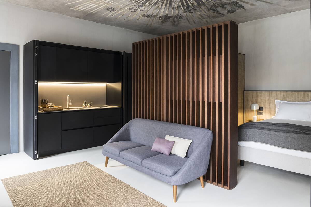 Quarto moderno com sofá cinzento, divisória de madeira, cama com cabeceira branca e guarda-roupa preto com prateleiras iluminadas.