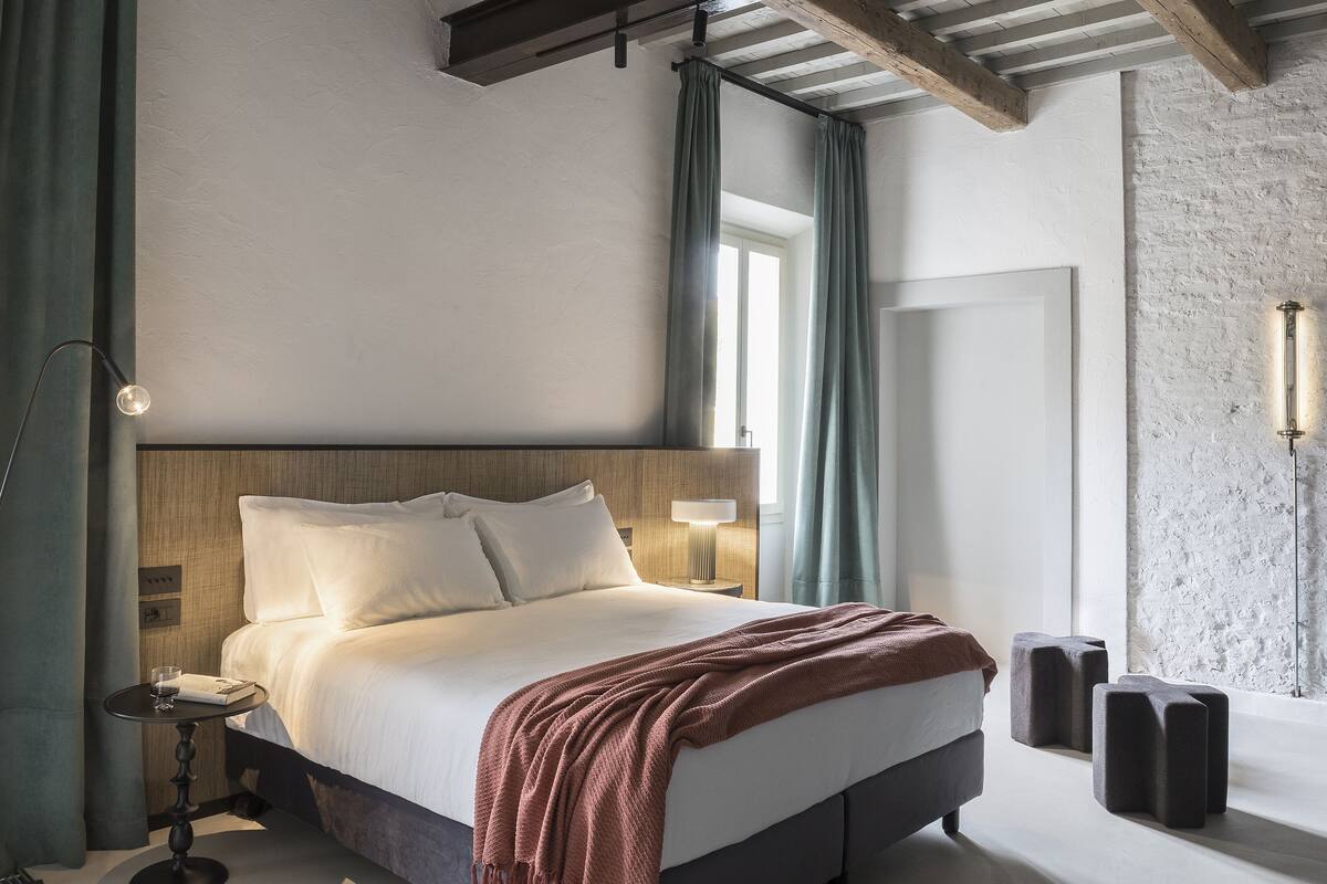 Quarto moderno com uma cama grande, mesa de cabeceira, luminária e vista para o exterior através de uma janela.