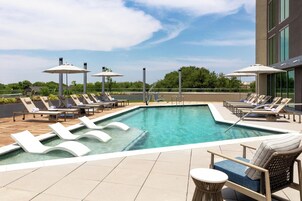Área de piscina no terraço com espreguiçadeiras e guarda-sóis, cercada por um edifício moderno.