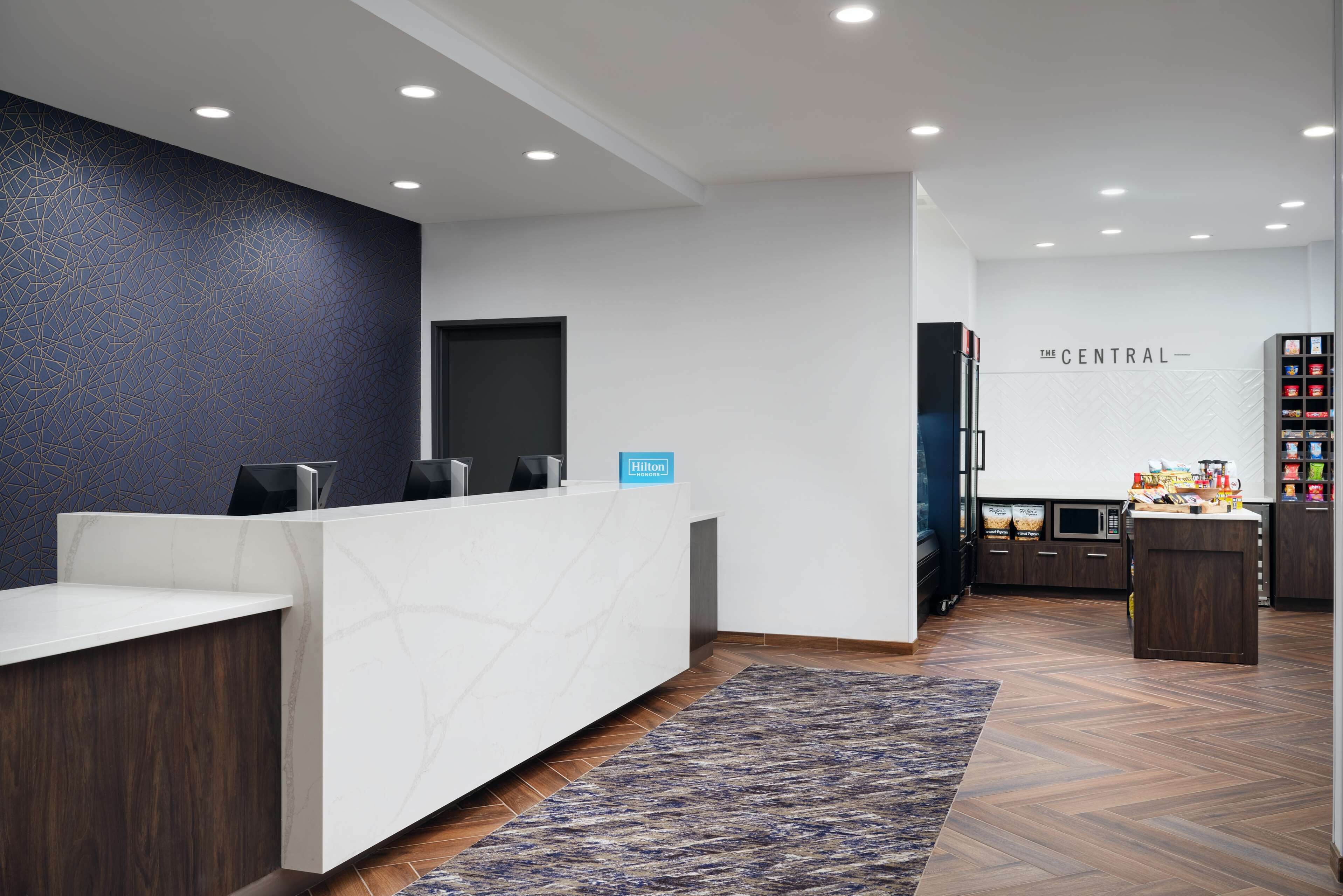 Uma área de recepção de hotel moderna, com um balcão de recepção branco, parede texturizada azul escura e uma cozinha com diversos eletrodomésticos e armários de armazenamento rotulada como “THE CENTRAL”.