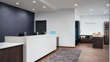 Uma área de recepção de hotel moderna, com um balcão de recepção branco, parede texturizada azul escura e uma cozinha com diversos eletrodomésticos e armários de armazenamento rotulada como “THE CENTRAL”.