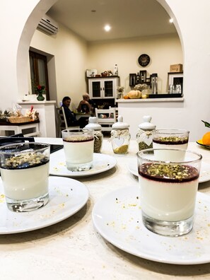 Uma área de refeições com pratos brancos e copos com sobremesas em camadas, uma cozinha ao fundo e um relógio na parede.