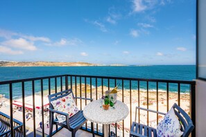 Varanda com mesa posta para dois, com vista para uma praia e um mar calmo, além de colinas ao longe.