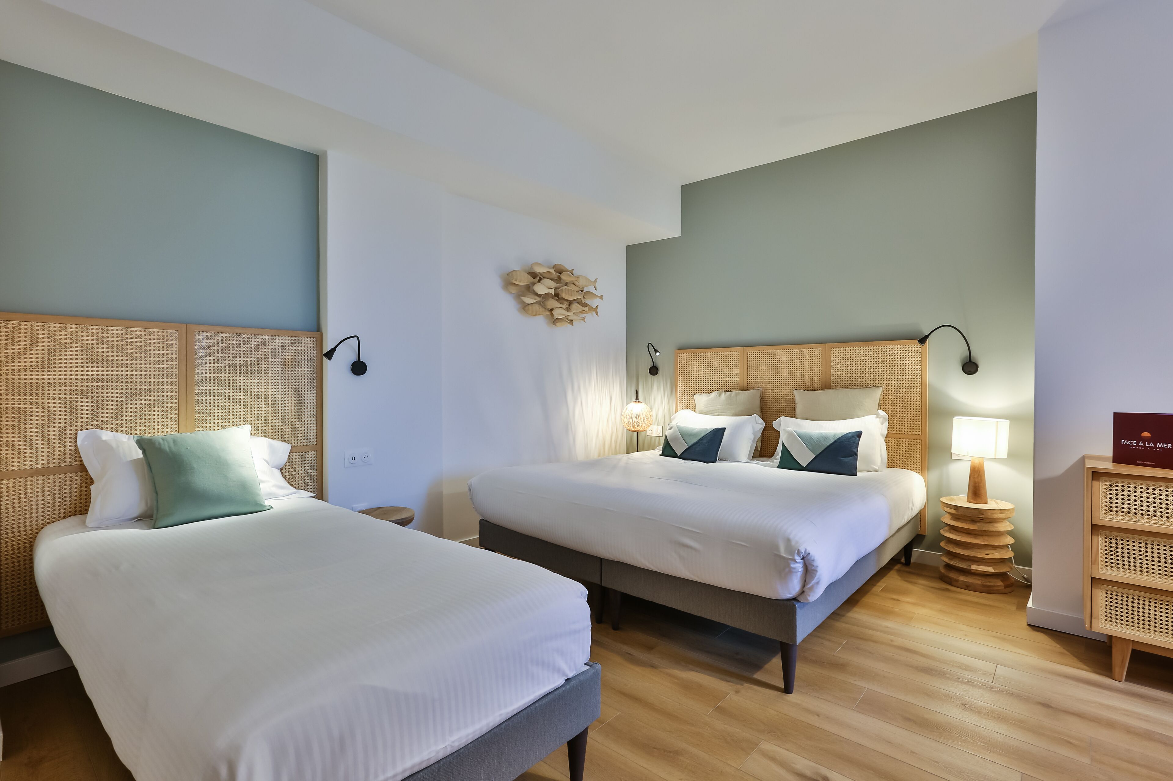 Quarto de hotel com duas camas, cabeceira de vime, luminária fixada na parede e mesa de cabeceira com abajur.