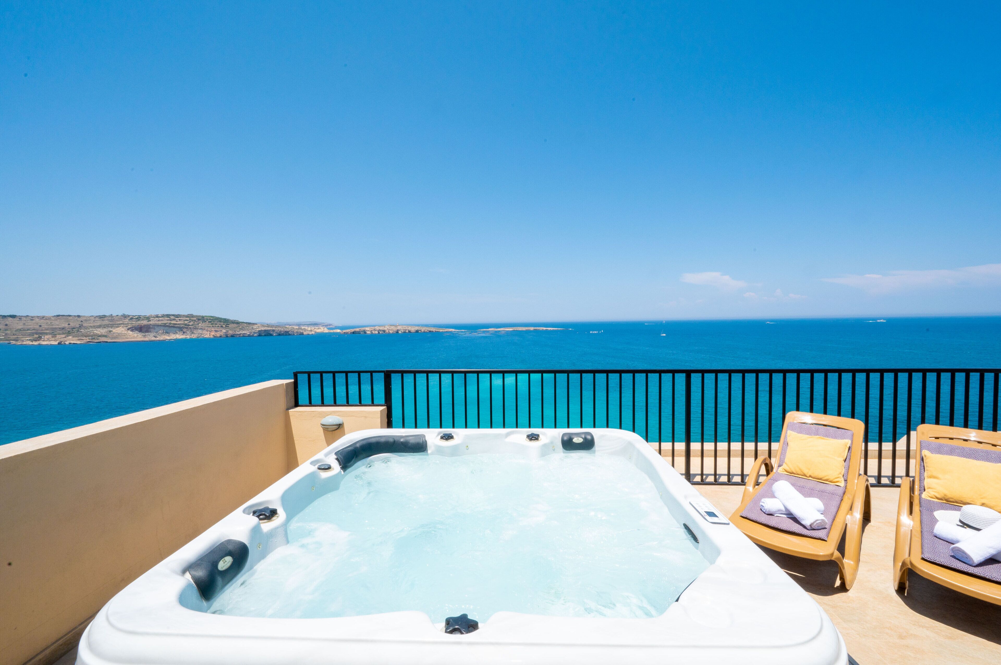 Uma banheira de hidromassagem com duas espreguiçadeiras numa varanda com vista para o mar azul e límpido.