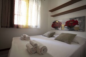 Cama bem arrumada com toalhas dobradas, um quadro com flores vermelhas e uma janela com cortinas leves.