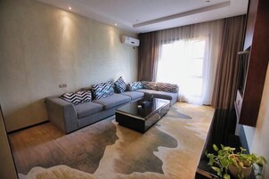 Uma sala de estar moderna com um sofá cinzento, almofadas estampadas, uma mesa de centro de vidro e uma janela ampla com cortinas.