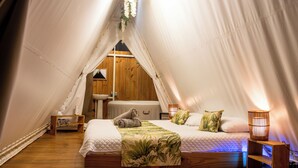 Um quarto aconchegante em estilo de tenda, com piso de madeira, uma cama com colcha estampada em folhas verdes e uma pequena mesa de cabeceira de madeira com abajur.