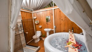 Uma casa de banho com decoração em madeira, cabine de duche em vidro, sanita, lavatório e hidromassagem com um tabuleiro de madeira que contém fruta e uma garrafa.