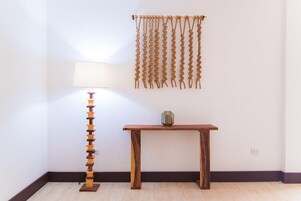 Canto de um quarto com piso de madeira, luminária com cúpula branca, mesa de madeira com um vaso e um objeto pendurado na parede.