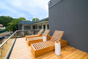Terraço de madeira com espreguiçadeiras e guarda-corpo de vidro com vista para a rua.