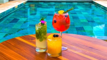 Três coquetéis sobre uma mesa de madeira junto a uma piscina com chão de azulejos.