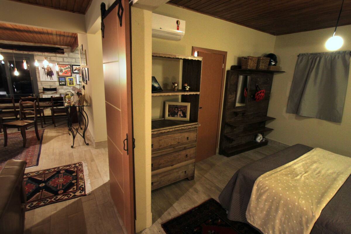 Quarto acolhedor com cama, cômoda, mesa pequena e cadeira. Há uma porta que dá para outro espaço e uma estante com objetos decorativos.