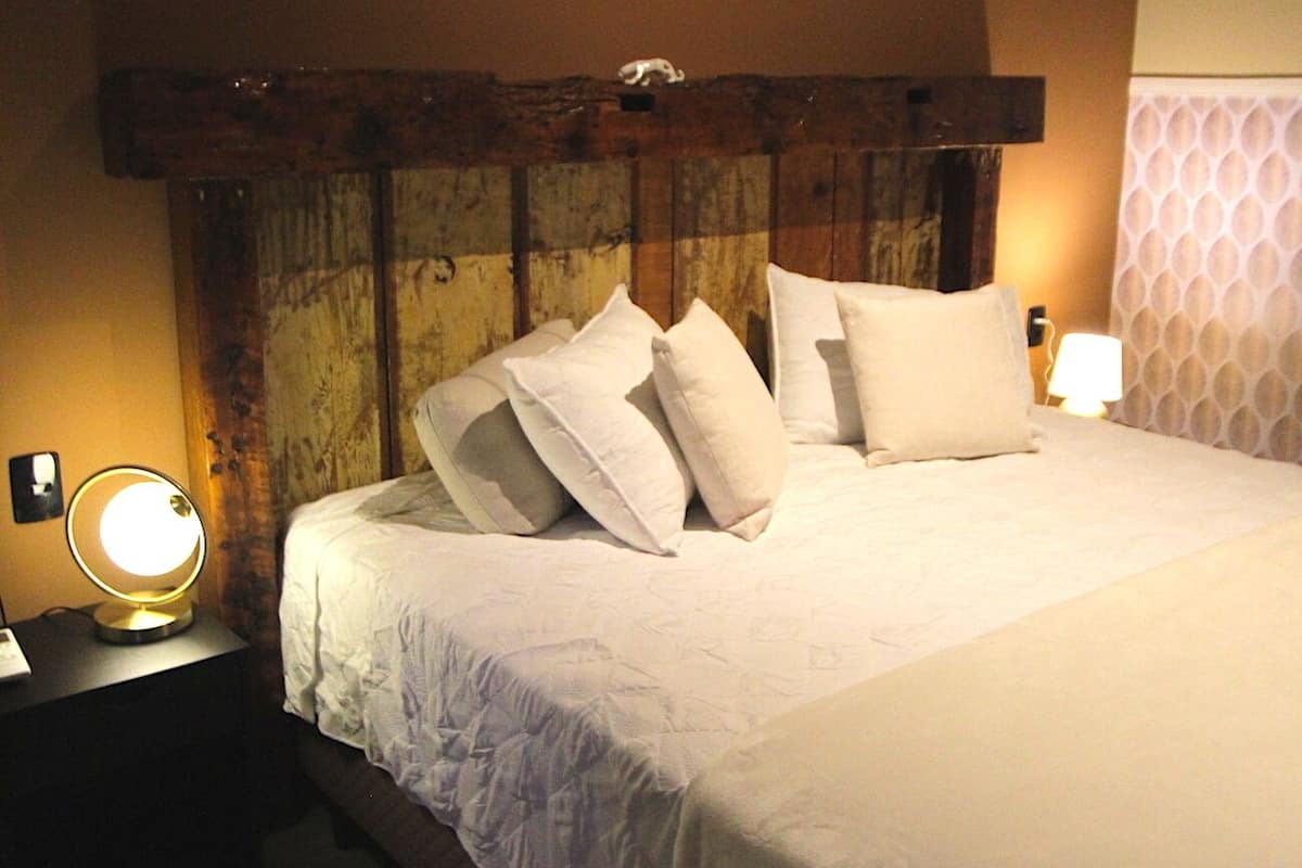 Uma cama bem arrumada com lençóis brancos e várias almofadas, um abajur e uma cabeceira de madeira.