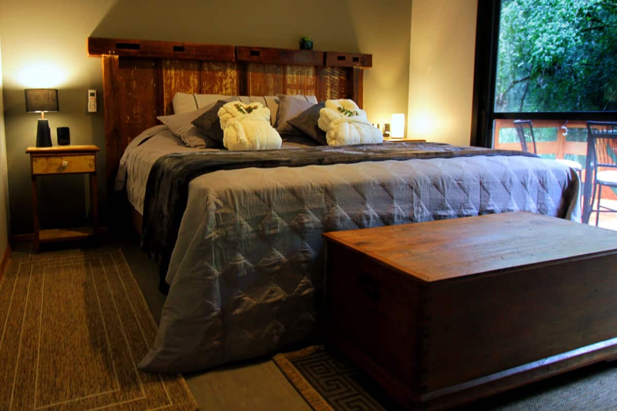 Quarto com duas camas, uma mesa de cabeceira, um banquinho de madeira e vista para o verde lá fora.