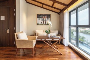 Quarto com piso de madeira, um sofá, uma cadeira, uma mesa com uma planta, um quadro na parede e uma janela com vista.
