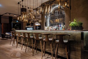 Uma área de bar com banquetas de madeira, luminárias pendentes e diversos frascos no balcão.