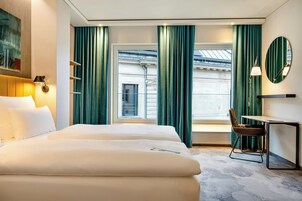 Quarto de hotel com uma cama grande, uma escrivaninha com cadeira, uma janela com cortinas e um candeeiro fixado na parede.