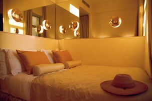 Quarto de hotel com uma cama grande, várias almofadas, um chapéu sobre a cama e um espelho refletindo a decoração do quarto.