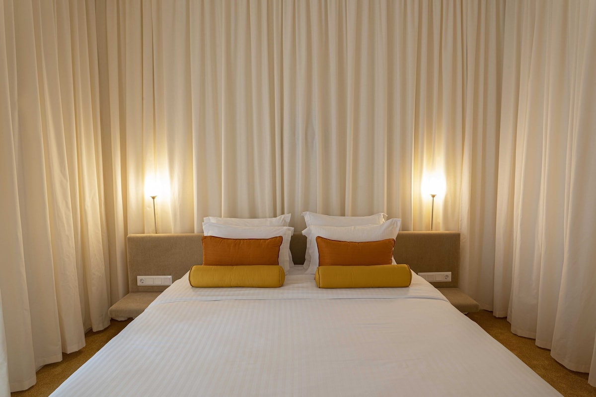 Quarto de hotel com uma cama grande, cortinas, duas luminárias de cabeceira e encosto de cama.