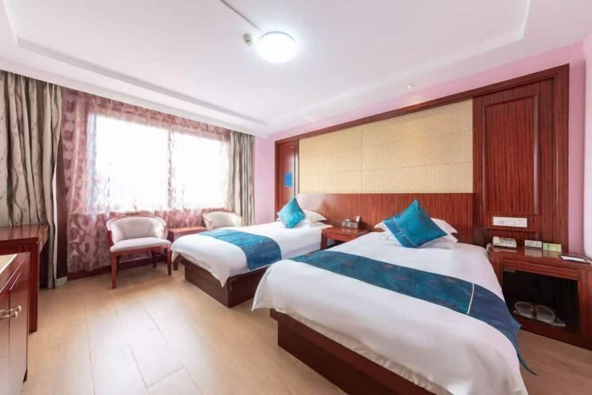 Quarto de hotel com duas camas, uma cadeira, uma escrivaninha e uma janela grande com cortinas.