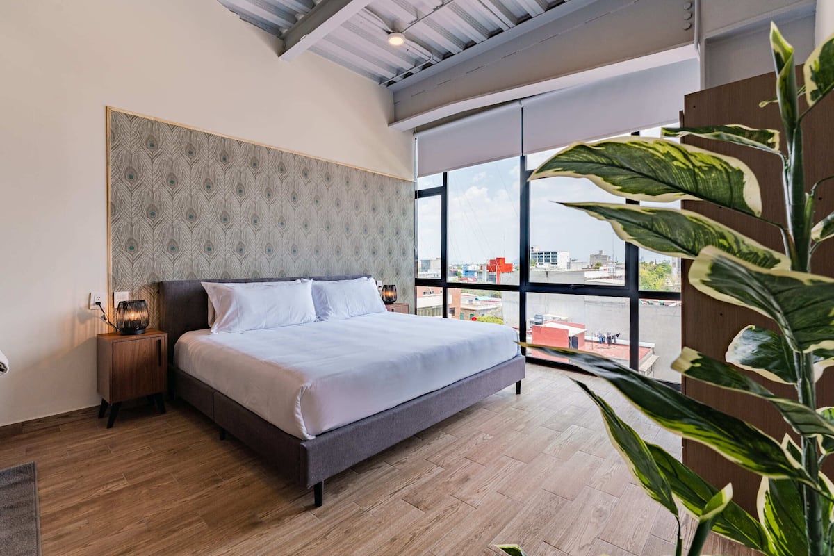 Quarto moderno com cama grande, cabeceira decorativa, mesa de cabeceira com abajur e vista através de janelas amplas.