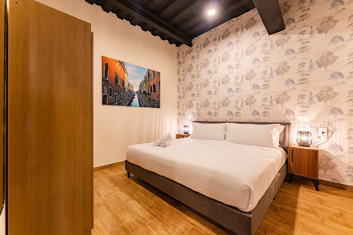 Cama bem arrumada num quarto com piso de madeira, um quadro de uma cena urbana na parede e papel de parede estampado.