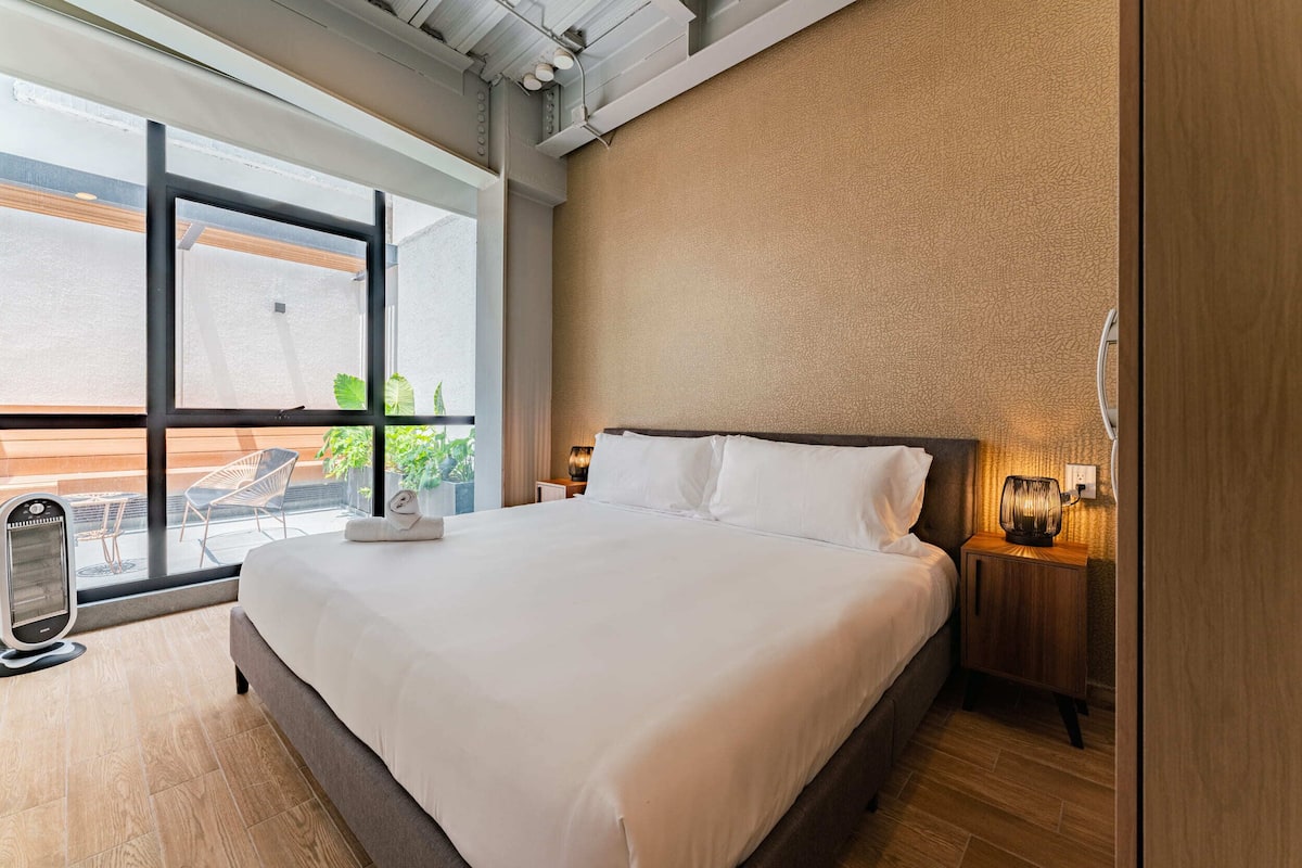 Quarto de hotel moderno com uma cama grande, mesa de cabeceira com abajur e vista para o exterior através de uma janela ampla.