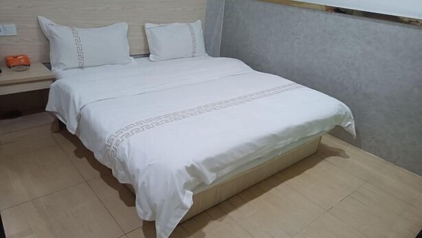 Cama bem arrumada com colcha branca e travesseiros, cabeceira de madeira e um objeto laranja pequeno na mesa de cabeceira.