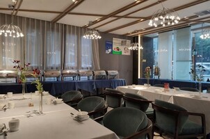 Uma sala de jantar com mesas postas, cadeiras e uma grande janela com cortinas.