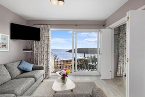 Uma sala de estar moderna com um sofá cinzento, uma televisão de ecrã plano, uma pequena mesa com um vaso de flores e uma varanda com vista para o mar.