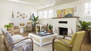 Uma sala de estar espaçosa com lareira branca, móveis de vime e diversos objetos decorativos.