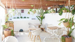 Um pátio externo com mobiliário de vime branco, vasos suspensos e vista para um edifício branco ao fundo.